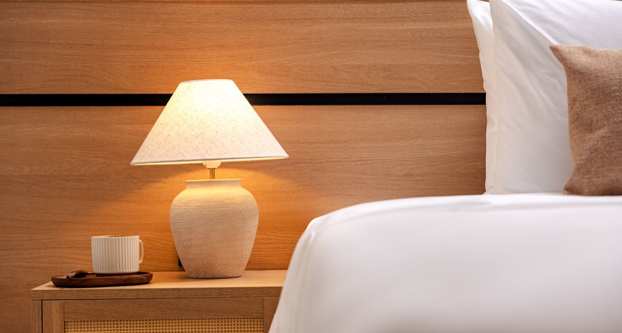 Uma cama bem arrumada com lençóis brancos e um travesseiro marrom, uma mesa de cabeceira com abajur, uma xícara branca e um tabuleiro de madeira.