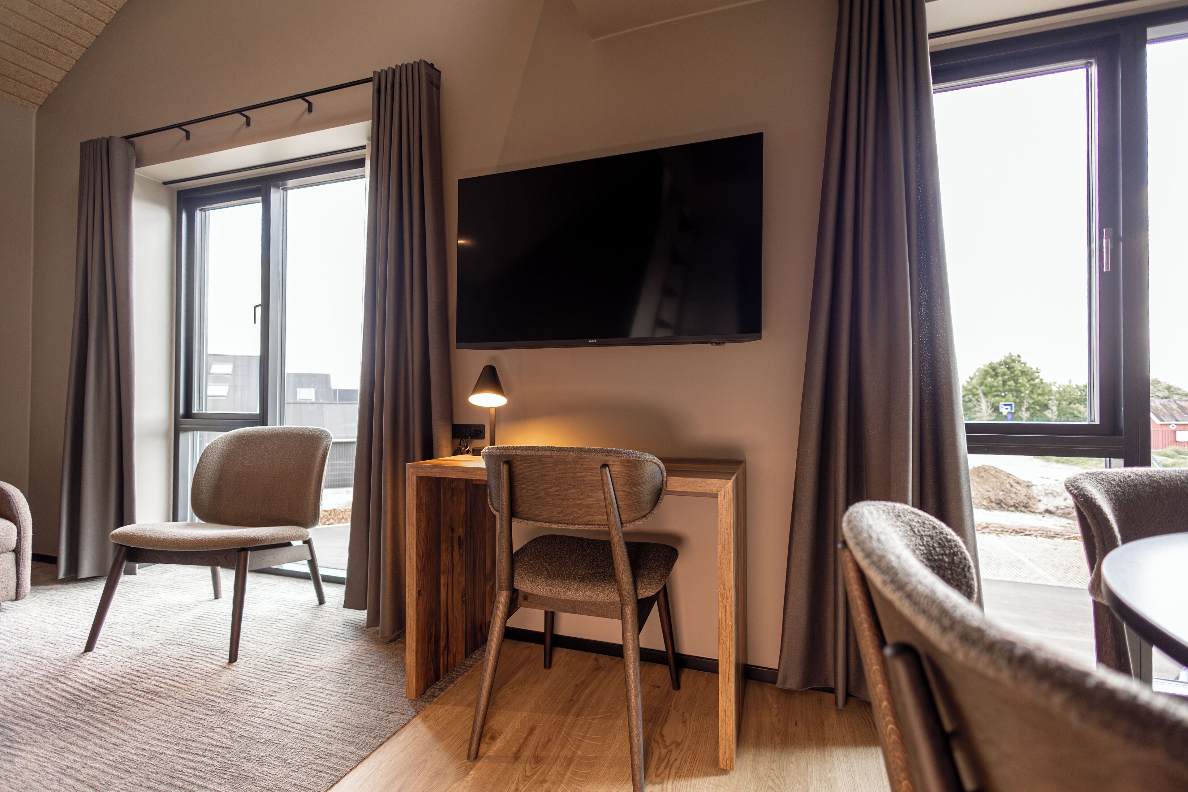 Quarto de hotel moderno com uma televisão grande fixada na parede, uma escrivaninha com uma cadeira e vista para o exterior através de amplas janelas.