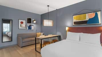 Quarto de hotel moderno com uma cama grande, um sofá, uma mesinha e uma escrivaninha. Há luminárias fixadas na parede e uma luminária no teto.