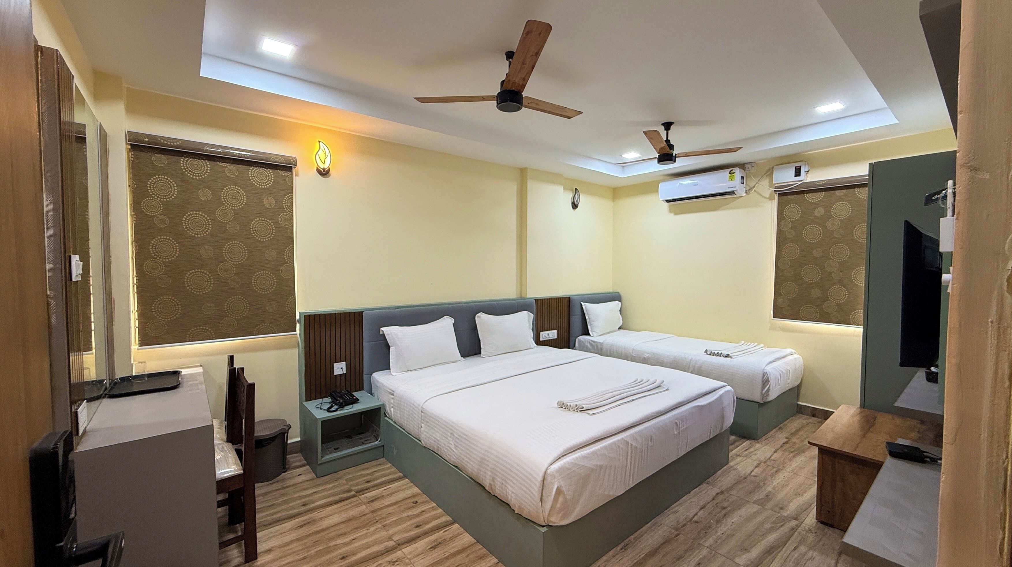 Quarto de hotel com duas camas, uma escrivaninha, uma cadeira, um espelho, um ventilador de teto e um ar condicionado.