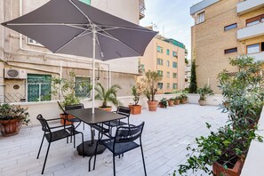Terraço no telhado com mesa e cadeiras, rodeado por plantas em vasos e um grande guarda-sol.