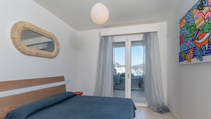Quarto com cabeceira de madeira, cama com colcha azul escura, espelho tecido na parede e pintura abstrata colorida.