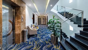 Um hall de hotel moderno com carpete estampado, um elevador de vidro, escada com guarda-corpos de vidro e um vaso de planta.