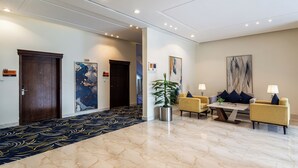 Um hall de hotel com piso de mármore, dois sofás, uma mesa de centro, uma planta em vaso e obras de arte nas paredes.