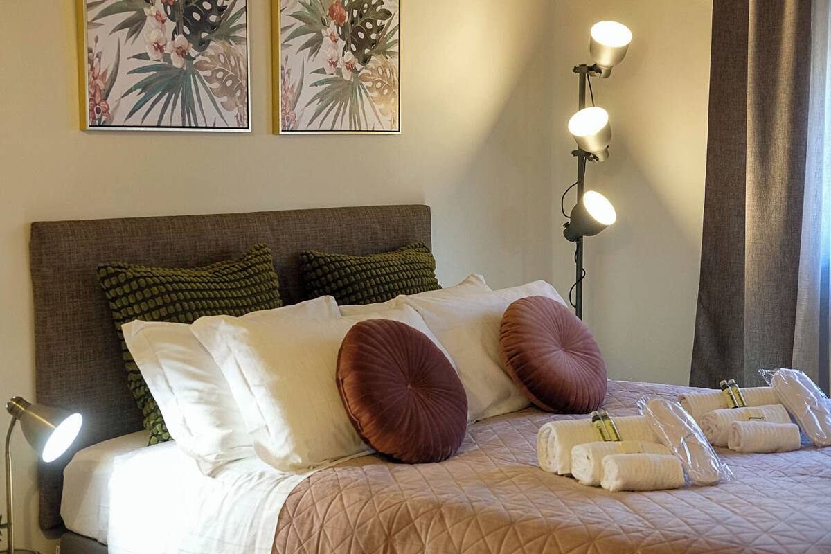 Cama bem arrumada com várias almofadas, abajur e luminária na parede.