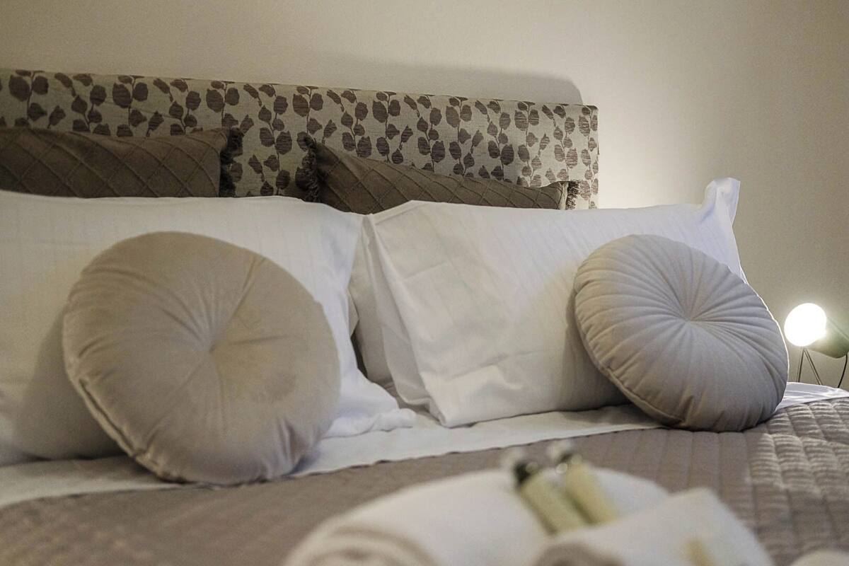 Uma cama bem arrumada com almofadas brancas e bege, um painel de cabeceira estampado e uma luminária de mesa.