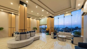 Um hall de hotel moderno com um sofá curvo, uma mesa de centro e vista para o horizonte urbano através de amplas janelas.