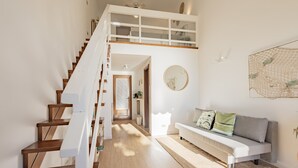 Uma sala de estar ampla com escada de madeira, um sof á branco e um espelho redondo na parede.