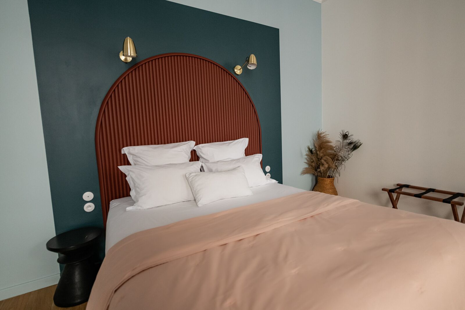 Uma cama bem arrumada com almofadas brancas e um cobertor cor de pêssego, uma cabeceira redonda de madeira e uma pequena mesa de cabeceira com um vaso de plantas secas.