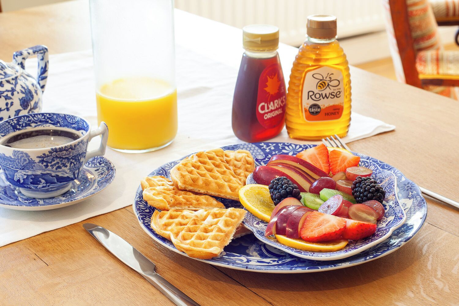 Uma mesa de café da manhã com waffles, frutas, mel e suco.