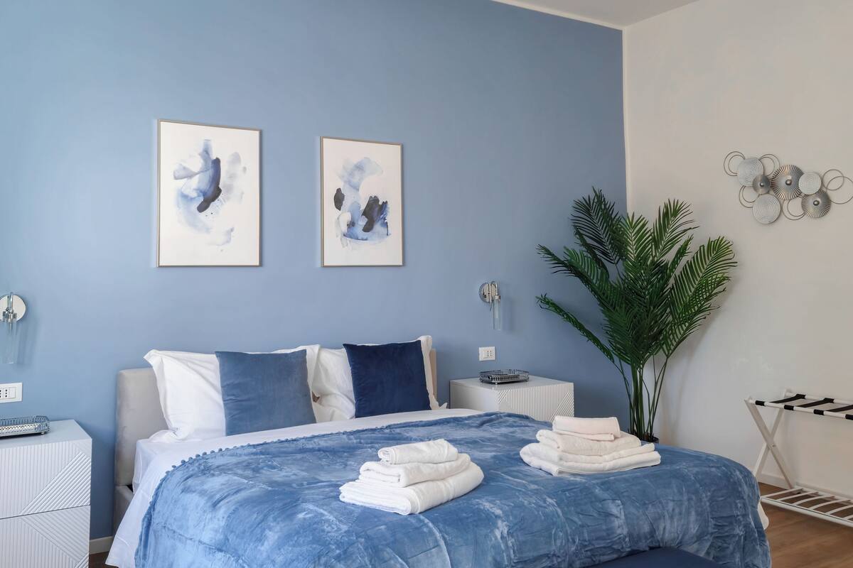 Quarto com cama, duas obras de arte emolduradas, um vaso de planta e um porta-toalhas fixado na parede.