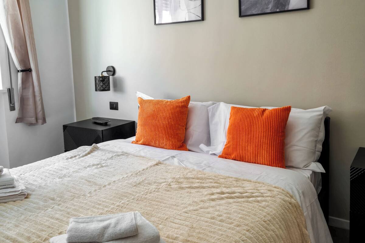 Quarto de hotel com cama, duas almofadas, mesa de cabeceira, luminária na parede e duas fotografias emolduradas.