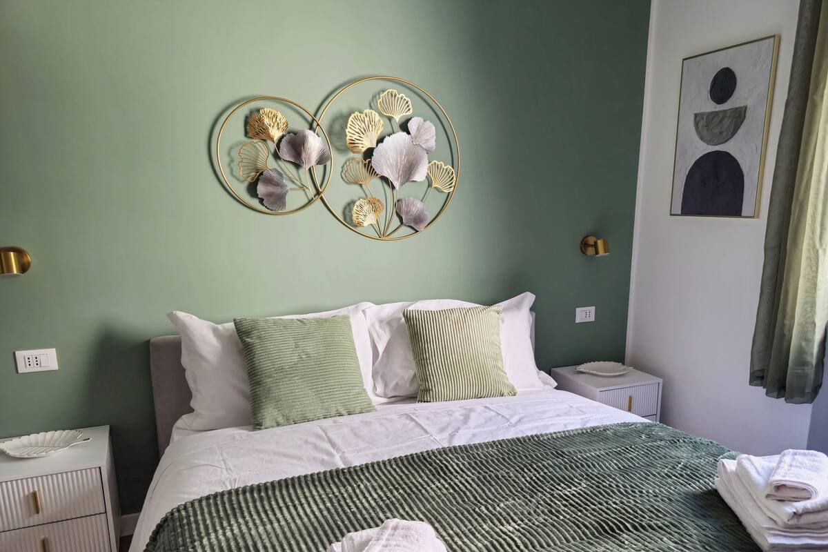 Cama bem arrumada com colcha verde texturizada, travesseiros brancos e toalhas dobradas, em parede verde com um detalhe metálico decorativo acima.