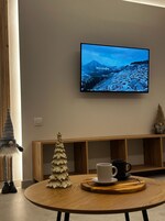 Sala de estar com mesa de madeira, uma pequena árvore de Natal, um gnomo e uma televisão exibindo uma paisagem nevada.