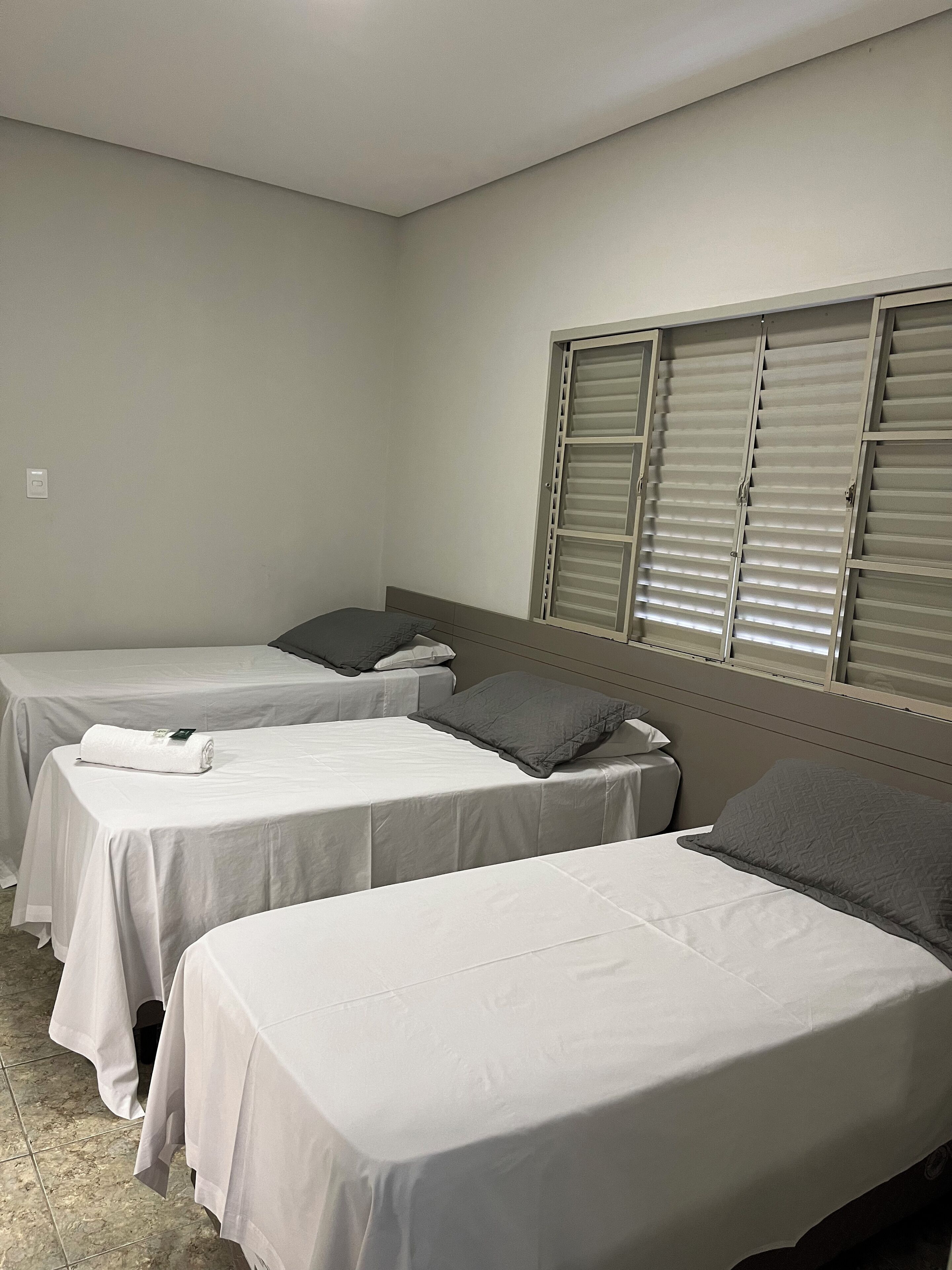 Quarto de hotel com três camas de solteiro, cada uma com uma colcha branca e um travesseiro. Há uma janela com persianas, um interruptor de luz na parede e piso de azulejos.
