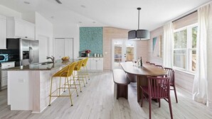 Uma cozinha moderna com armários brancos, uma ilha central com banquetas, uma área de jantar com mesa de madeira e cadeiras vermelhas, e uma janela grande com cortinas.
