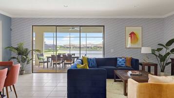 Uma sala de estar ampla com um sofá grande, uma mesa de centro e um espaço de jantar com cadeiras e mesa. Há uma porta de vidro deslizante que dá para um pátio externo com vista para casas e uma montanha.