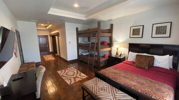 Quarto de hotel com beliches, uma escrivaninha, uma cadeira, cama com colcha estampada e mesa de cabeceira com abajur.