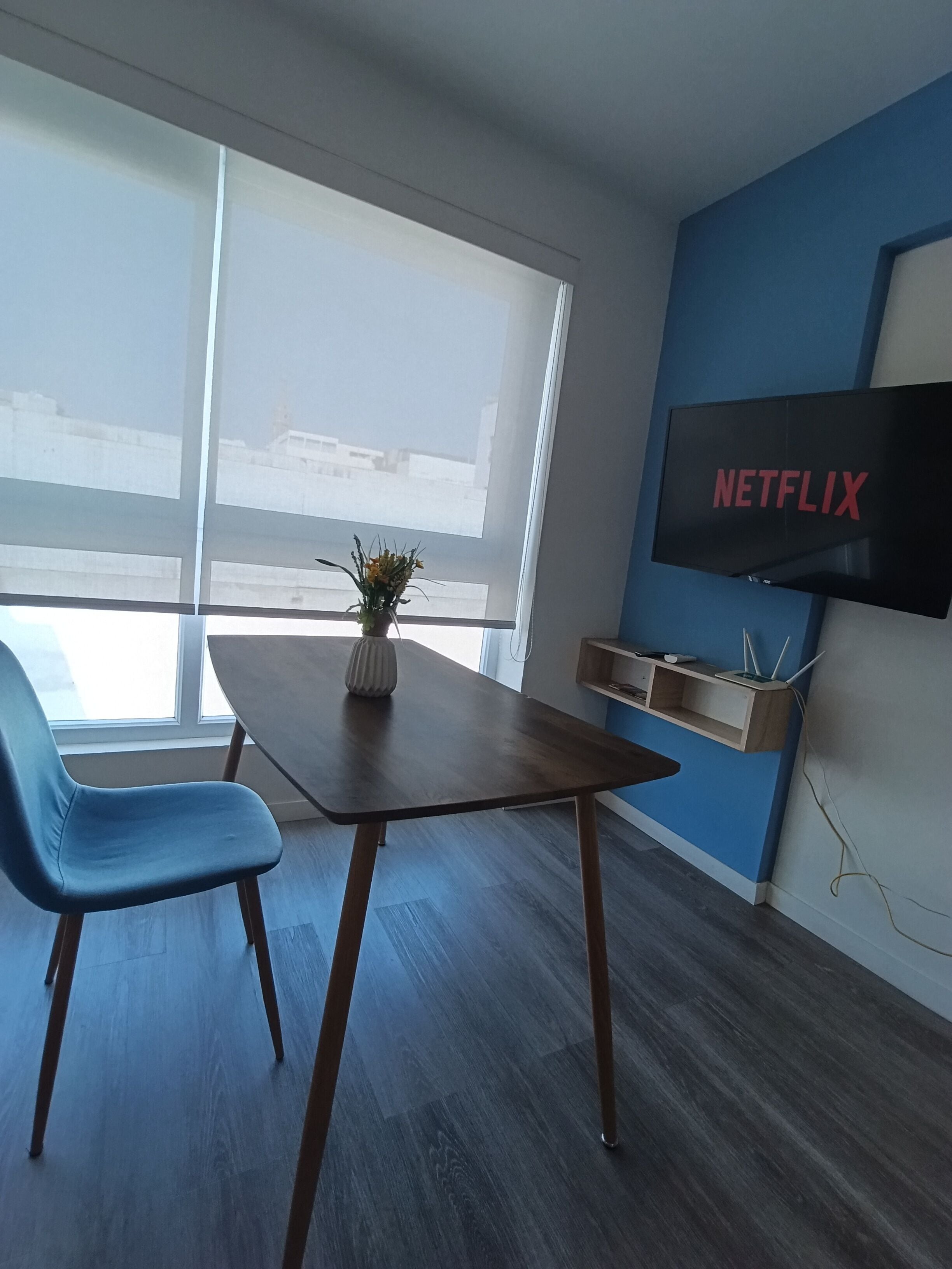 Quarto moderno com mesa de madeira, uma cadeira azul, planta em vaso, TV fixada na parede e vista através de grandes janelas.