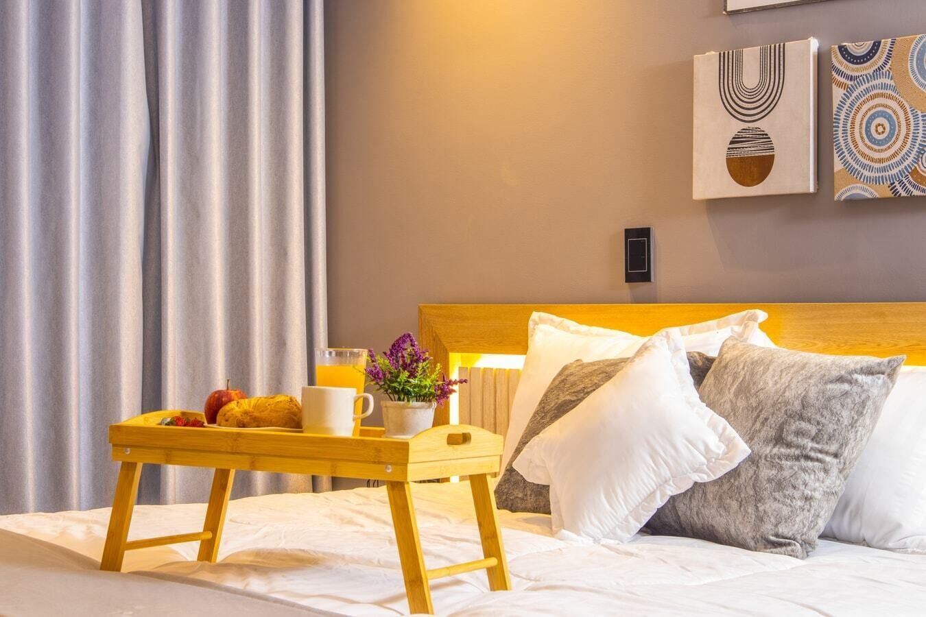 Quarto de hotel com uma cama, mesa de cabeceira de madeira com itens de café da manhã e uma obra de arte na parede.