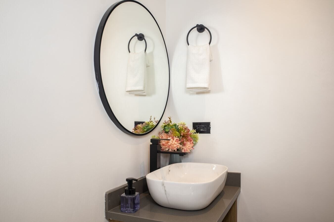 Um banheiro com um espelho oval grande, dois porta-toalhas, uma pia branca, um dispensador de sabonete e um vaso com flores.