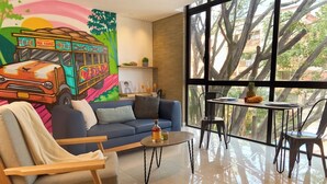 Uma sala de estar moderna com um sofá azul, uma cadeira de madeira, uma mesa de centro e uma grande janela com vista para árvores e edifícios.