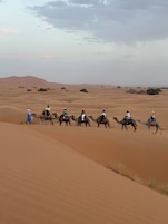 Um grupo de pessoas a cavalgamento de camelos atravessa uma paisagem desértica com dunas de areia.