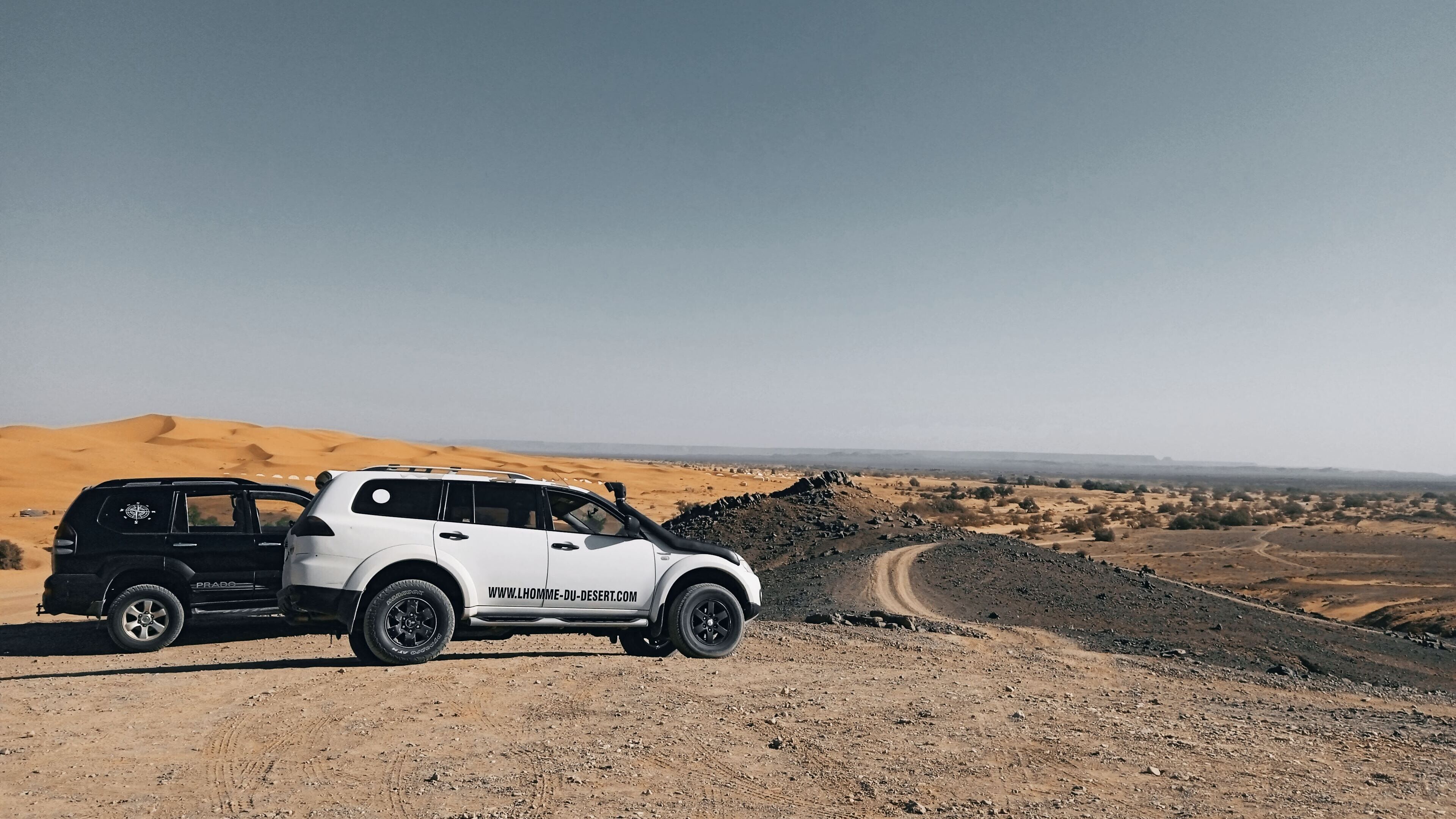 Um SUV branco estacionado em uma estrada no deserto, com dunas ao fundo.