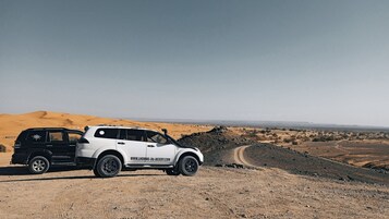 Um SUV branco estacionado em uma estrada no deserto, com dunas ao fundo.