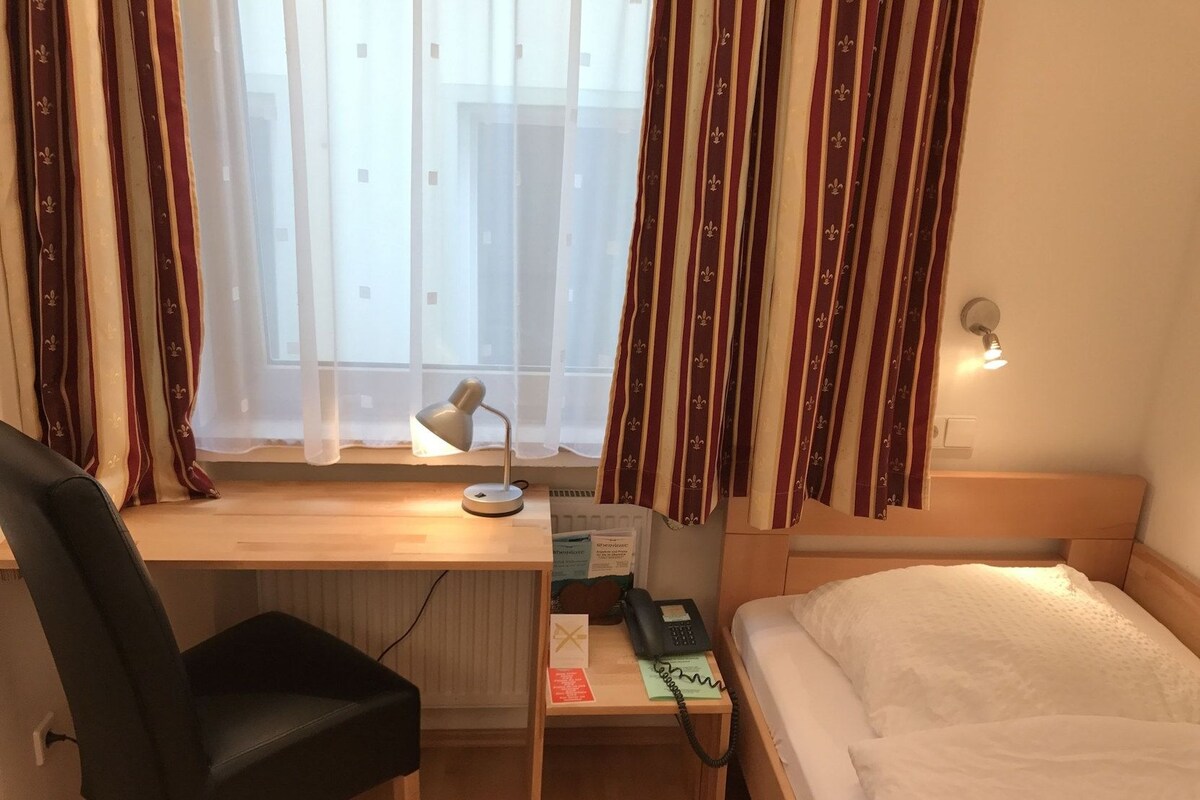 Quarto de hotel com escrivaninha, cadeira, cama e janela com cortinas.