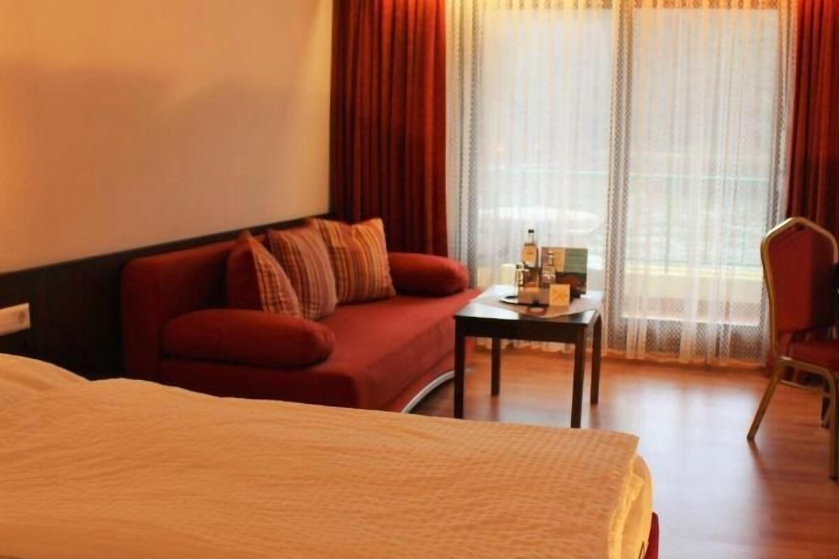 Quarto de hotel com cama, sofá vermelho, mesa pequena com uma garrafa, cadeira e escrivaninha.