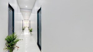Um corredor comprido e estreito, com paredes brancas e portas pretas, decorado com plantas em vasos.