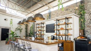 Uma área de bar com um balcão comprido, várias cadeiras, luminárias pendentes, prateleiras com garrafas e um forno a pizza.