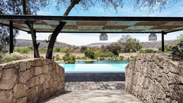 Área de piscina com paredes de pedra, coberta por uma pérgola, cercada por paisagem natural e colinas.