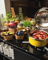 Uma bancada de cozinha com diversos alimentos, incluindo potes de picles, um recipiente com bacon e uma tigela de ervas.