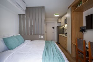 Quarto com cama, mesa de cabeceira, escrivaninha com cadeira, cozinha compacta e banheiro.