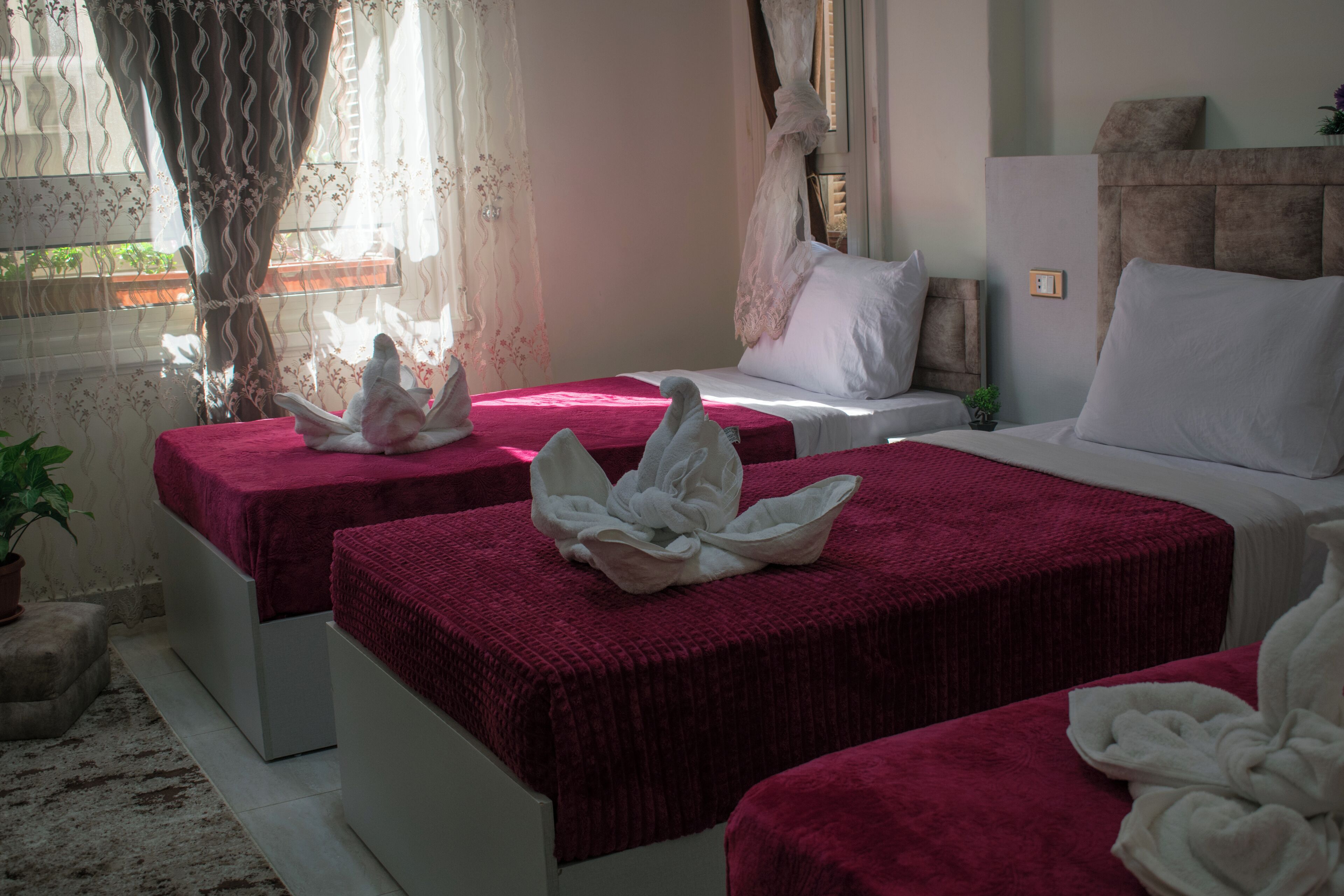 Quarto de hotel com duas camas, cada uma com colcha vermelha e almofadas brancas. Há uma janela com cortinas, uma mesa de cabeceira com uma planta e um aplique na parede.