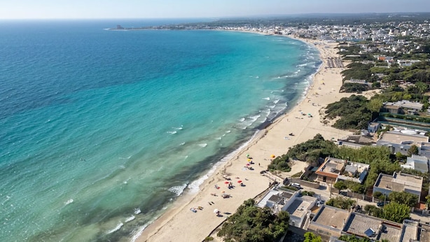 Vista aérea de uma vila costeira com praia, edifícios e mar azul cristalino.