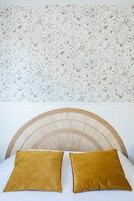 Cama com duas almofadas mostarda e cabeceira de tecido, em frente a uma parede com padrão floral.