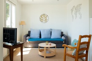Uma sala de estar com um sofá cinza, uma mesa de centro redonda de madeira, uma cadeira de madeira, uma TV sobre uma mesa e um mapa-múndi na parede.