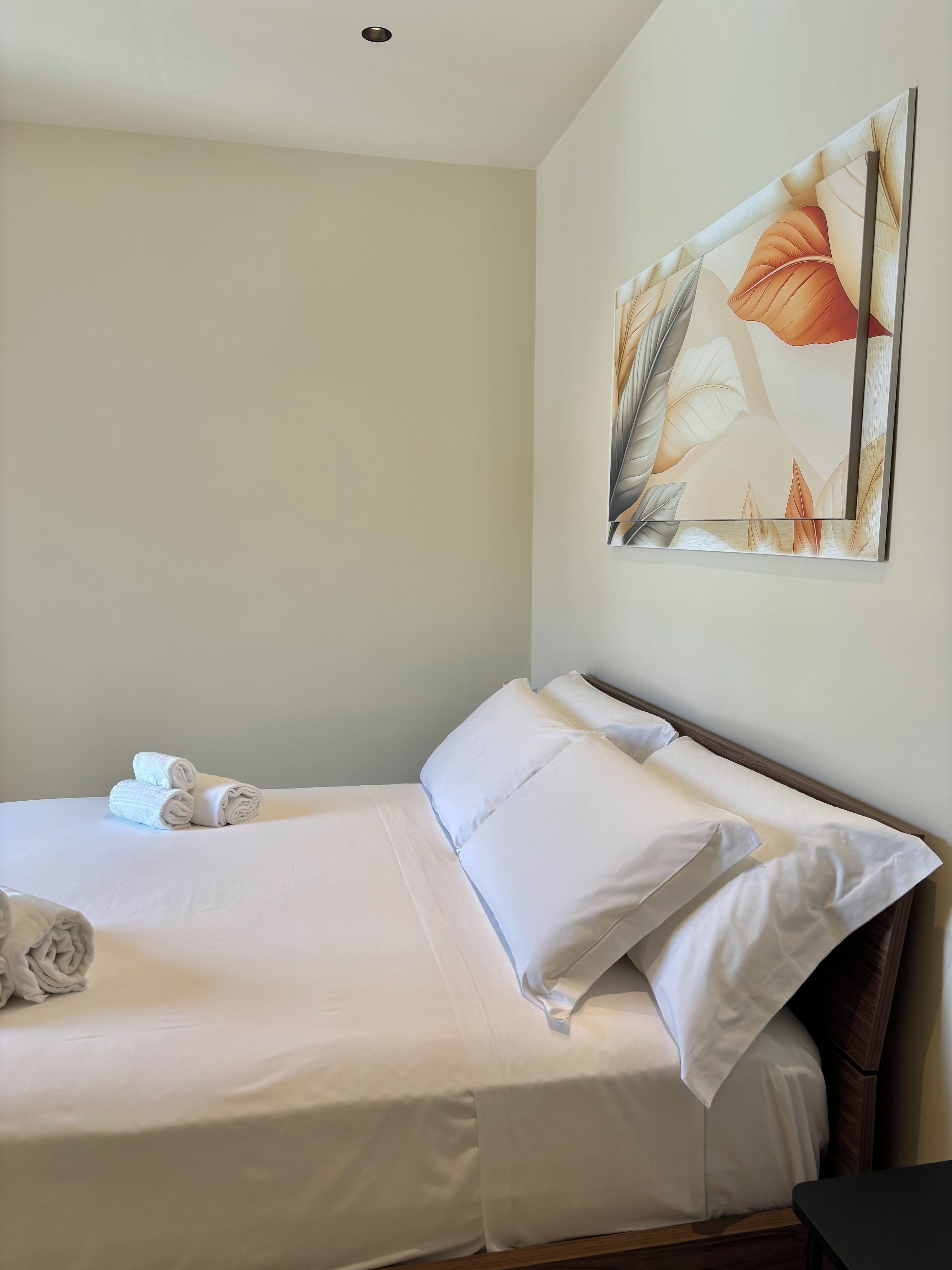 Cama bem arrumada com lençóis e almofadas brancos, uma obra de arte na parede retratando folhas, e cabeceira de madeira.