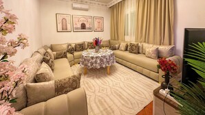 Uma sala de estar com um sofá bege, uma mesa de centro redonda com toalha de mesa florida, um lustre e uma grande janela com cortinas.