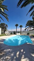 Piscina com espreguiçadeiras, palmeiras e um céu azul límpido.
