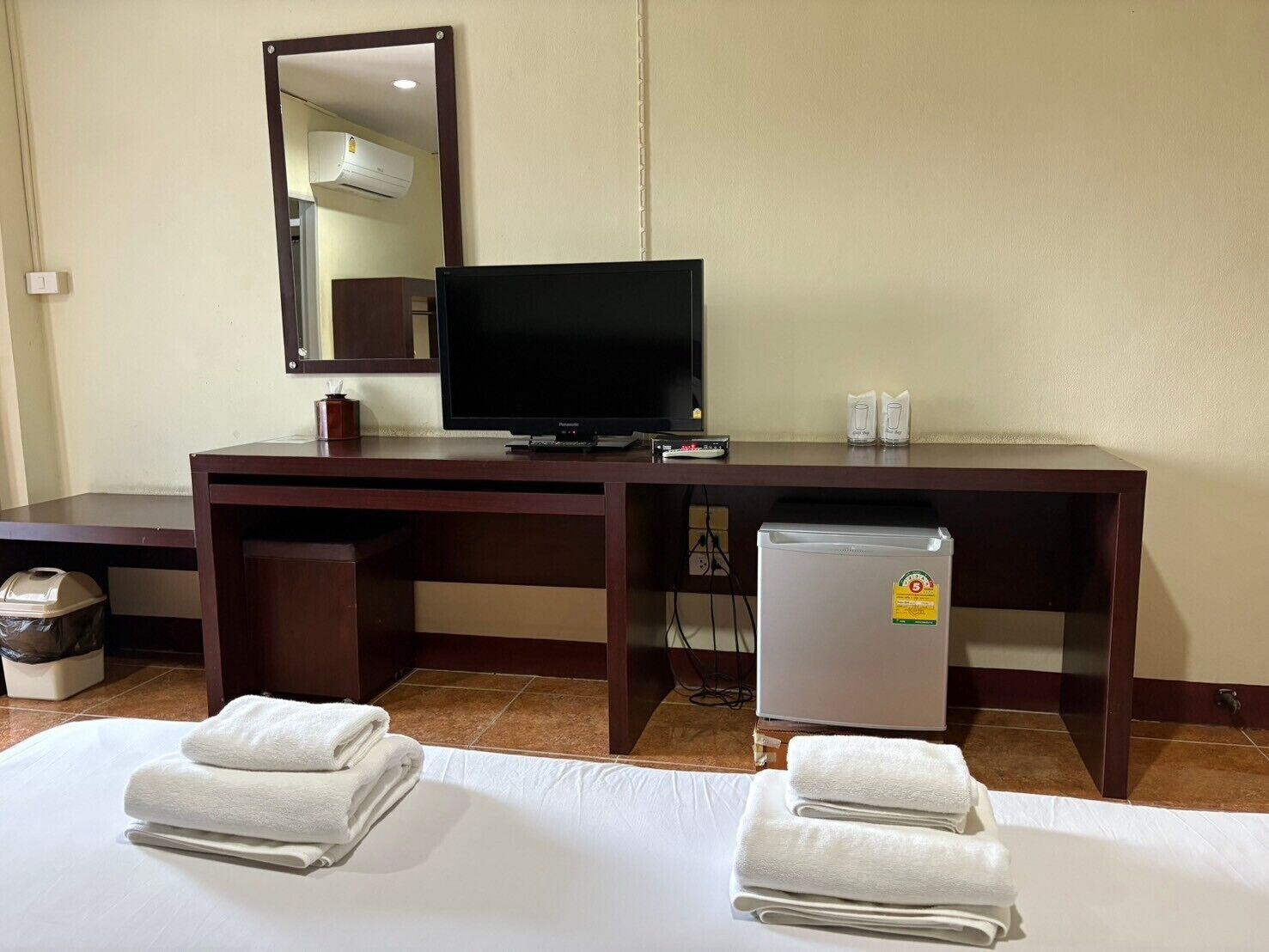 Quarto de hotel com cama, escrivaninha, televisão, mini-geladeira e lixeira.