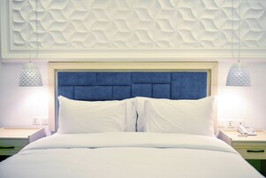 Quarto de hotel com cabeceira azul, roupa de cama branca, duas mesinhas de cabeceira com luminárias e teto texturizado.