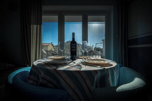 Uma mesa de jantar com uma garrafa de vinho, copos e pratos, com vista para prédios e um céu limpo através de uma janela.