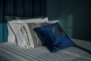 Uma cama com roupa de cama listrada, um travesseiro branco, um travesseiro estampado e um travesseiro azul com emblema dourado.