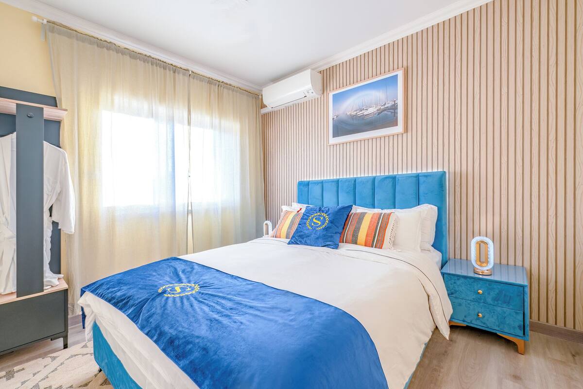 Quarto com cama, mesa de cabeceira, guarda-roupa e janela com cortinas.