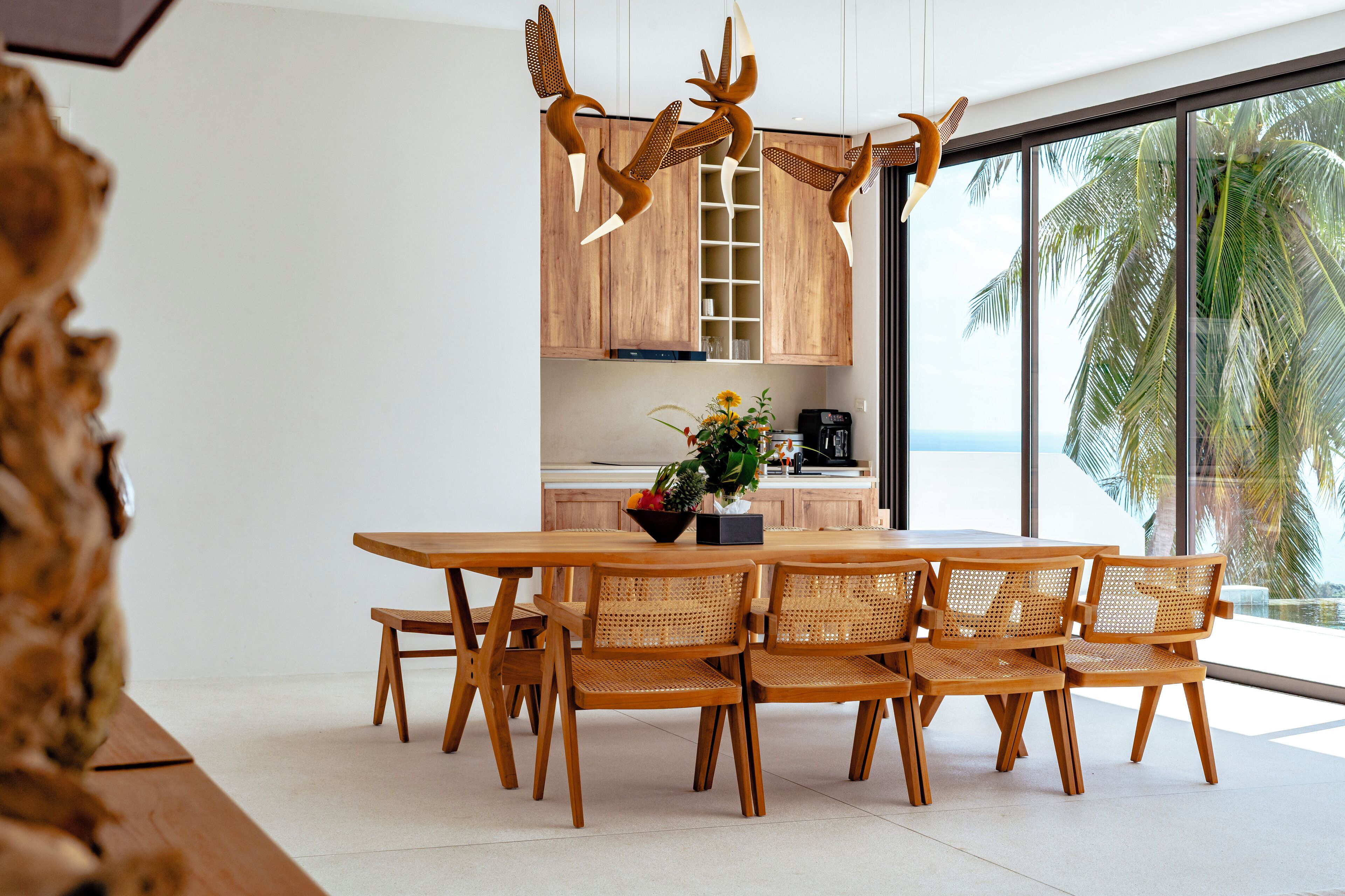 Uma cozinha moderna com armários de madeira, uma mesa de jantar com cadeiras de vime e vista para palmeiras através de grandes janelas.