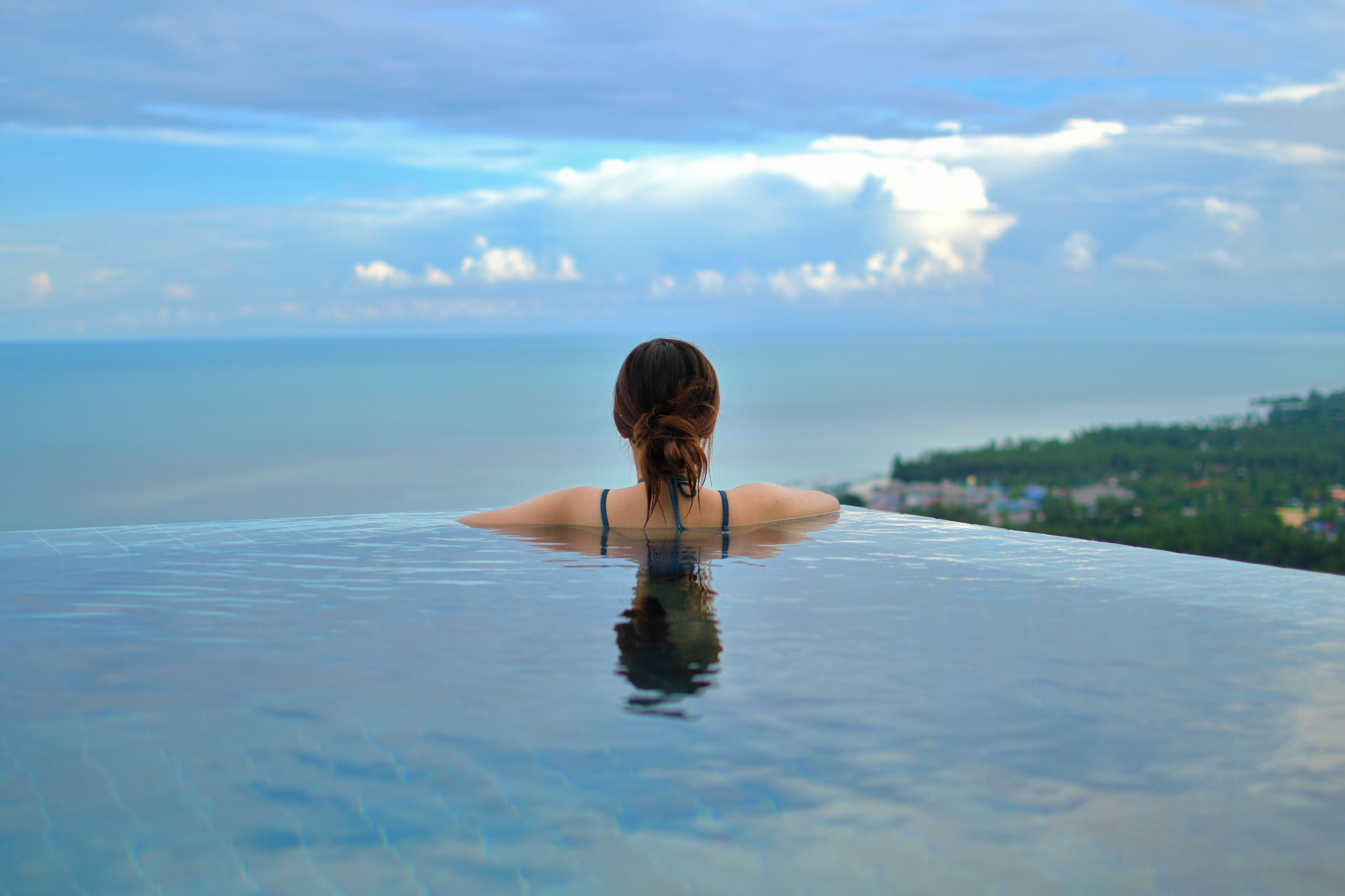 Uma mulher de biquíni preto sentada na borda de uma piscina infinita, com vista para o mar e uma linha de costa distante.